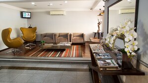 Um lobby moderno com um sofá, duas poltronas amarelas, uma mesa de centro, revistas, um espelho e um arranjo de flores.