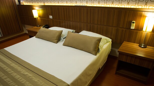 Quarto de hotel com uma cama grande, duas mesas de cabeceira e um telefone fixado na parede.