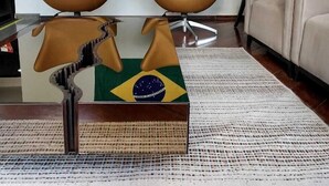 Uma sala de estar com duas poltronas de couro marrom, uma mesa de centro de vidro com o desenho da bandeira do Brasil, um tapete estampado e um sofá.