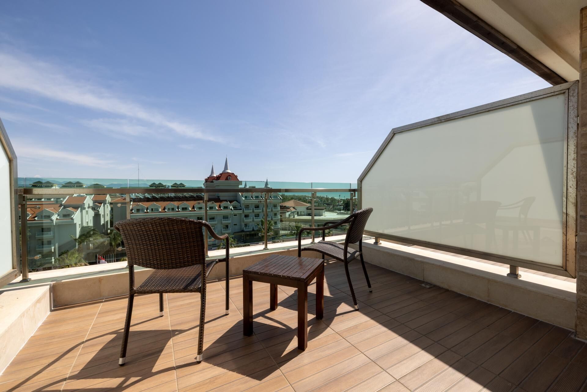 Uma varanda com duas cadeiras de vime e uma pequena mesa, com vista para um horizonte urbano com edifícios e uma torre ao longe.