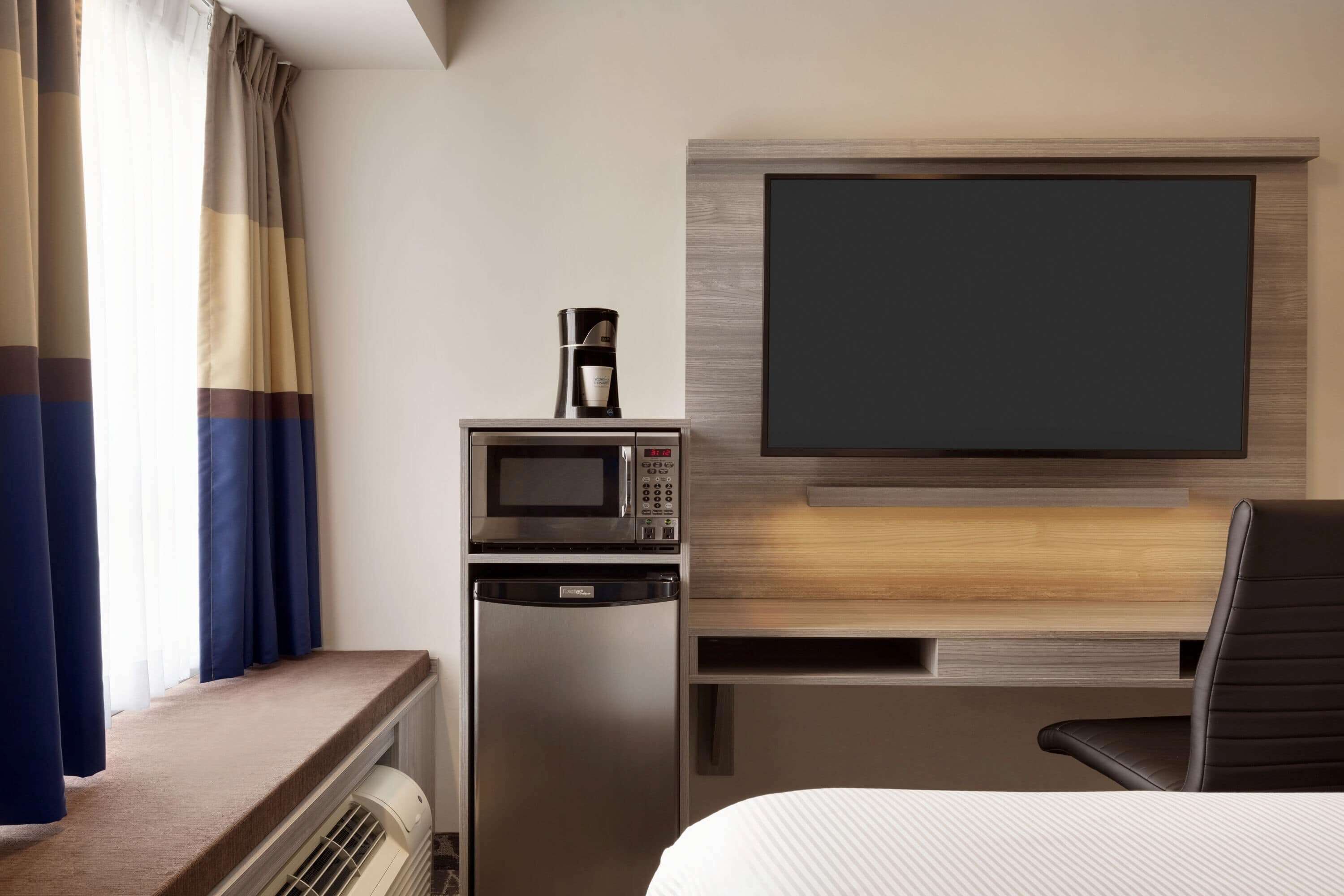 Quarto de hotel com cama, escrivaninha com computador, micro-ondas, máquina de café e TV de tela plana na parede.