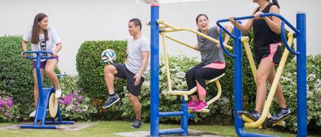 Um grupo de pessoas se exercitando em equipamentos de ginástica ao ar livre, em um jardim.