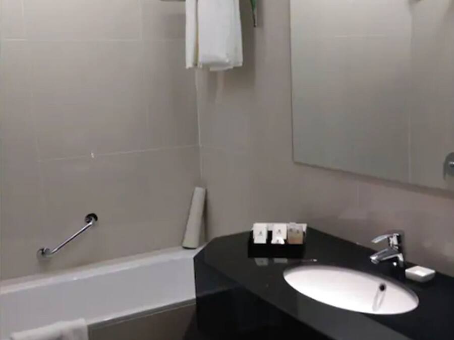Uma casa de banho moderna com bancada preta, lavatório branco e um espelho grande. Há um porta-toalhas com uma toalha dobrada e um rolo de papel higiénico.