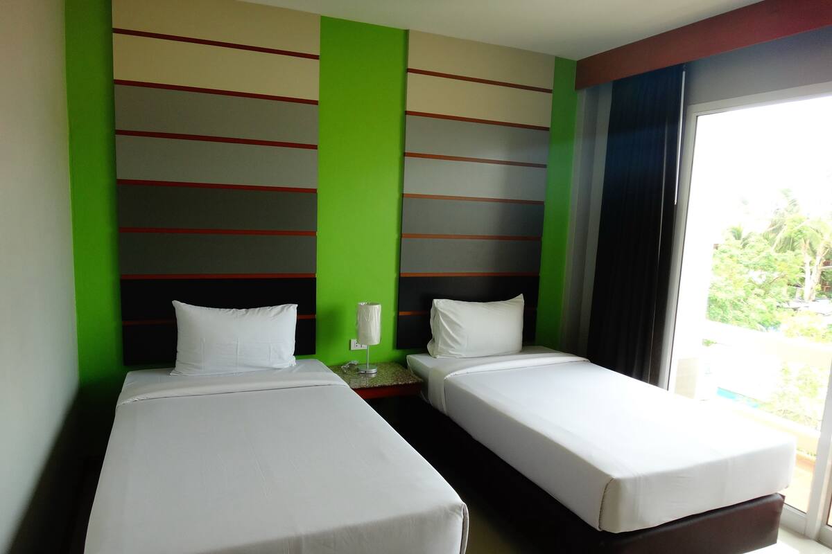 Quarto de hotel com duas camas, uma mesa de cabeceira e janela com vista para o verde lá fora.