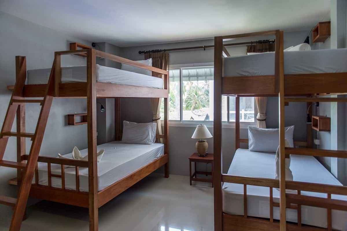 Quarto com duas beliches, uma pequena mesa de cabeceira com abajur e uma janela com cortinas.