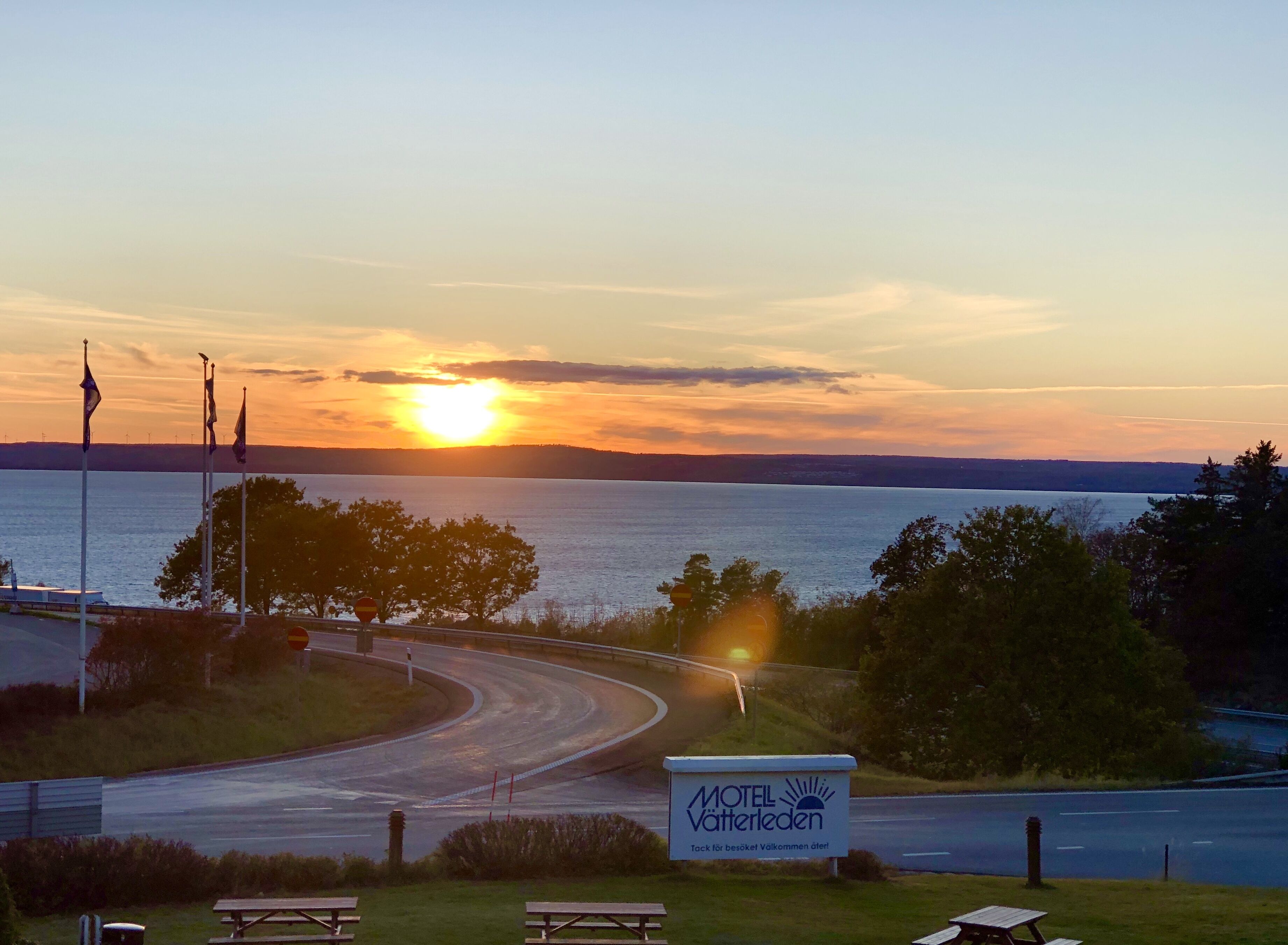 Vista do pôr do sol com uma estrada curvando-se para a direita, uma placa indicando 'Motel Vätterleden' e um corpo d'água ao fundo.