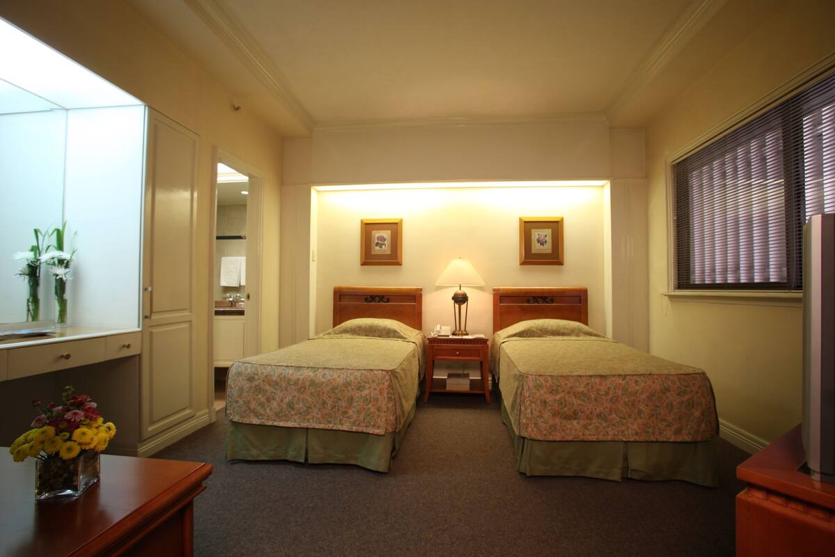 Quarto de hotel com duas camas, mesa de cabeceira, luminária, espelho e janela com persianas.