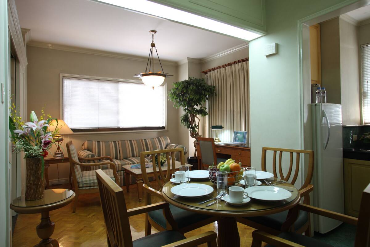 Uma sala de jantar com uma mesa redonda posta para quatro pessoas, rodeada por cadeiras de madeira. Há um sofá com almofadas listradas ao fundo, uma planta em vaso e uma janela com persianas.