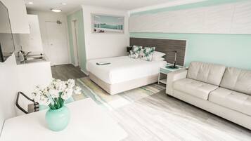 Quarto de hotel moderno com uma cama, um sofá, uma mesinha com um vaso de flores e uma pintura de uma cena de praia.