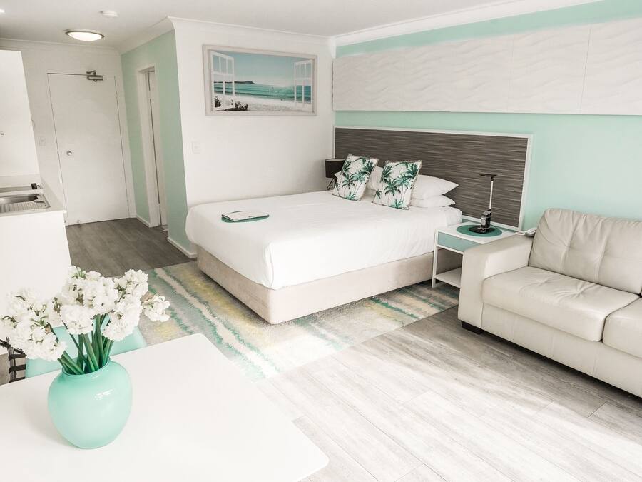Quarto de hotel moderno com uma cama, um sofá, uma mesinha com um vaso de flores e uma pintura de uma cena de praia.