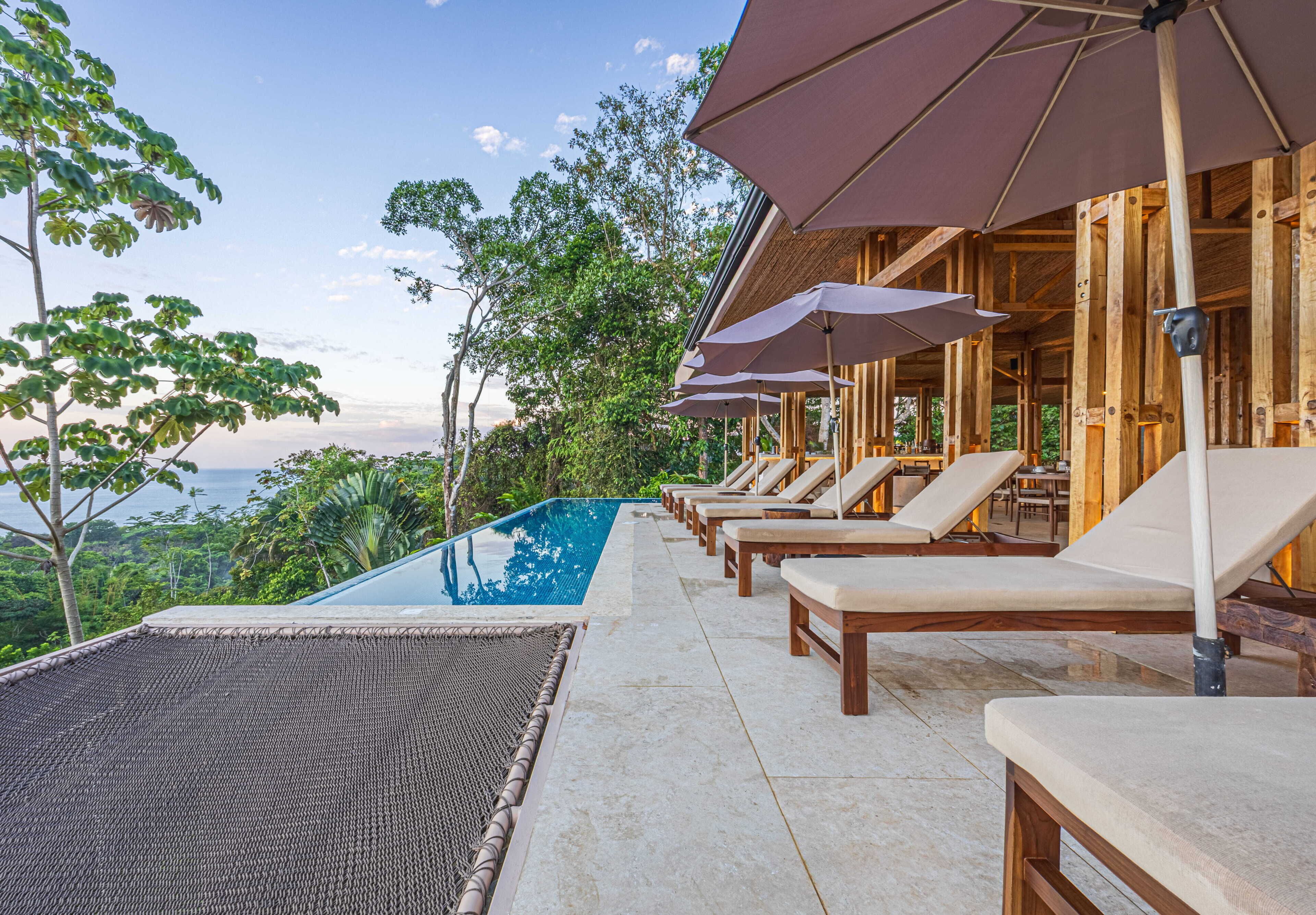 Área à beira da piscina com espreguiçadeiras e guarda-sóis, com vista para a água e vegetação.