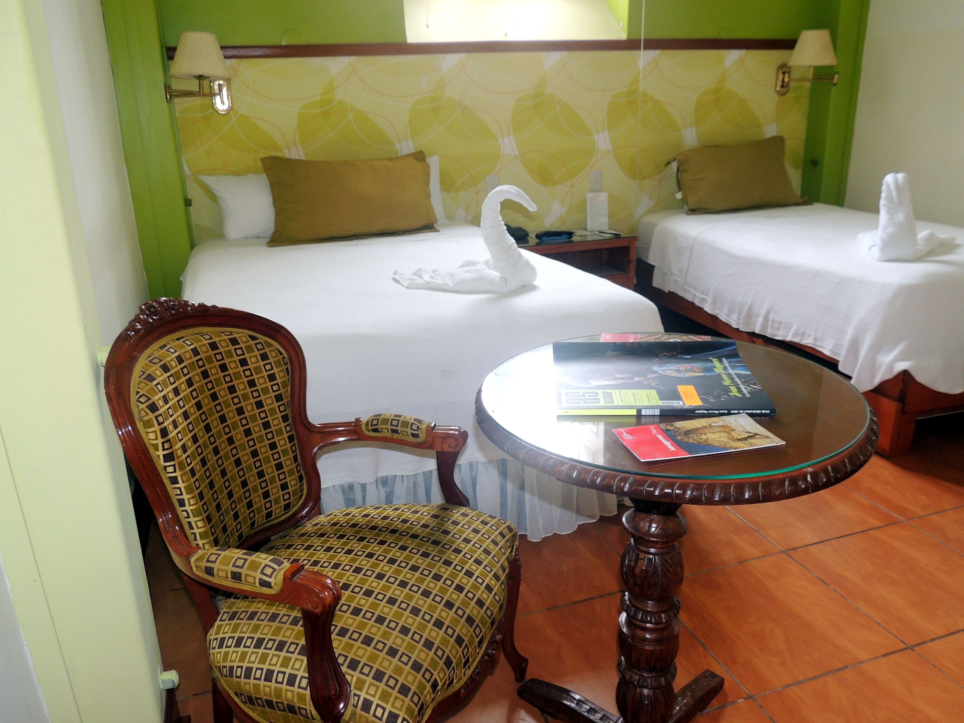 Quarto de hotel com duas camas, uma mesa redonda com livros e uma poltrona estampada.