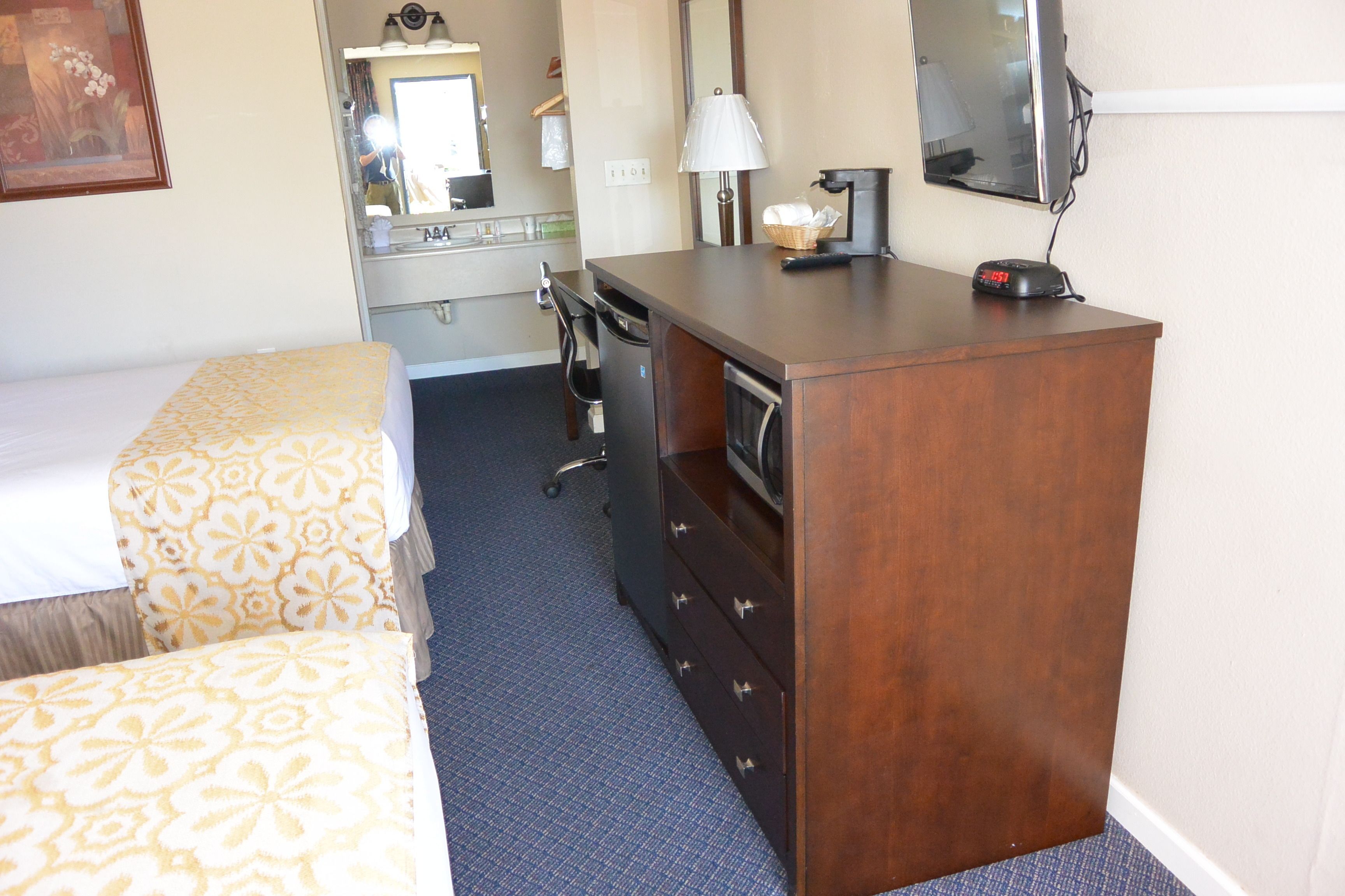 Quarto de hotel com duas camas, uma cômoda de madeira, uma escrivaninha com luminária, um aparelho de café e um espelho.