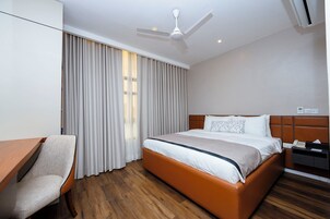 Quarto de hotel com cama, escrivaninha, cadeira, ventoinha no teto e janela com cortinas.