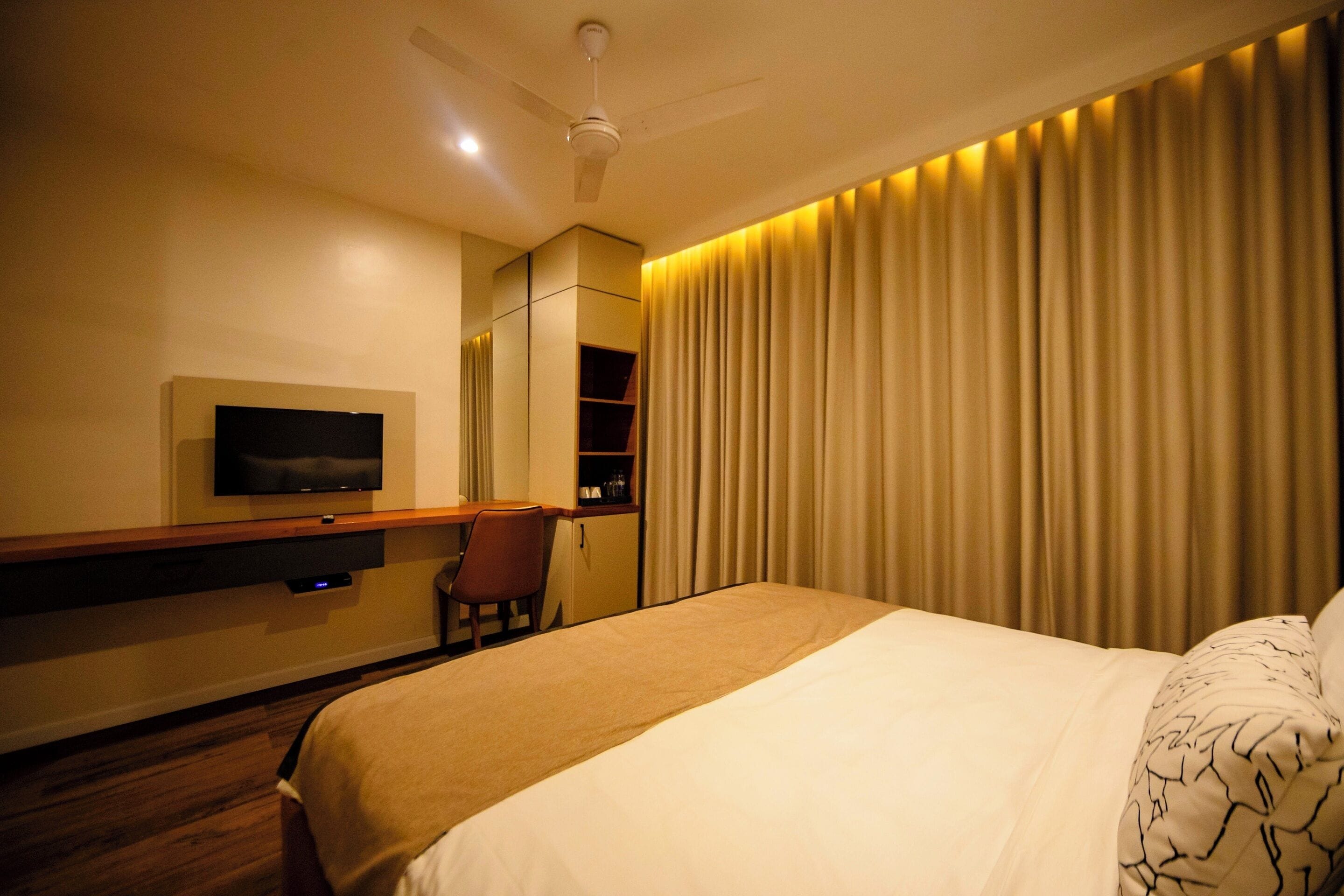 Quarto de hotel com uma cama, uma escrivaninha com televisão, uma cadeira e uma janela grande com cortinas.