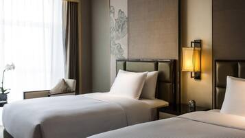 Quarto de hotel com duas camas, mesa de cabeceira com abajur, uma obra de arte na parede e janela com cortinas.