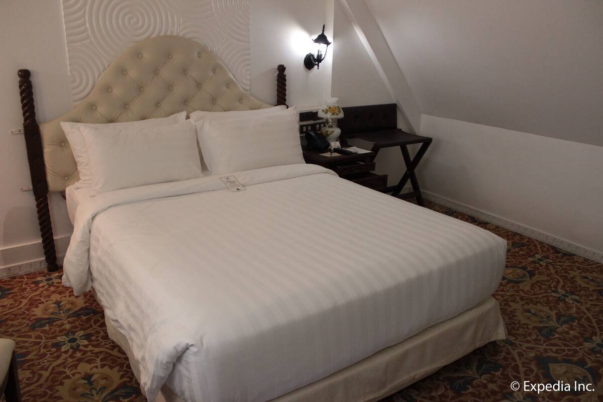 Uma cama bem arrumada com cabeceira estofada, mesa de cabeceira com abajur e tapete estampado.