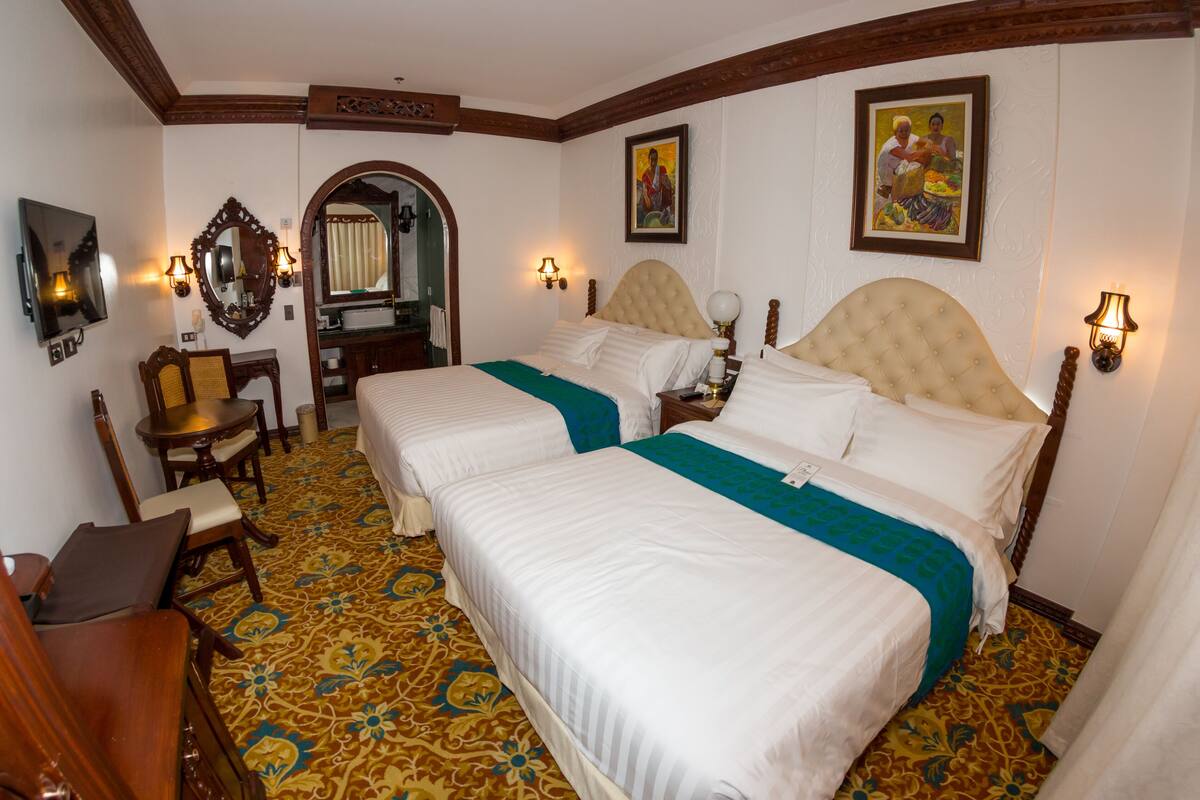 Quarto de hotel com duas camas, área de estar com uma cadeira e mesa, espelho e obras de arte nas paredes.
