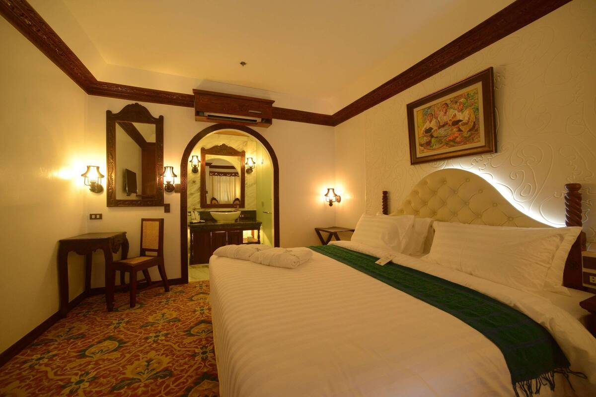 Quarto de hotel com uma cama grande, uma cadeira, toucador com espelho e um quadro na parede.