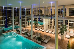 Piscina interior em vários níveis com áreas de estar, paredes de vidro e luzes pendentes azuis.