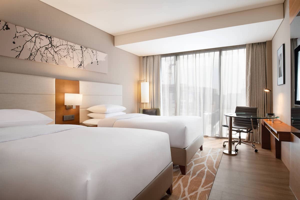 Quarto de hotel com duas camas, uma escrivaninha e uma cadeira. Há uma janela grande com cortinas, uma luminária na parede e um quadro sobre a cama.