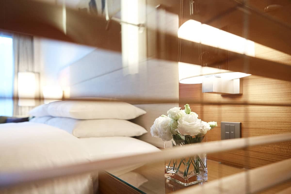 Quarto de hotel com cama, almofadas brancas, mesa de cabeceira com um vaso de flores e luminária moderna.