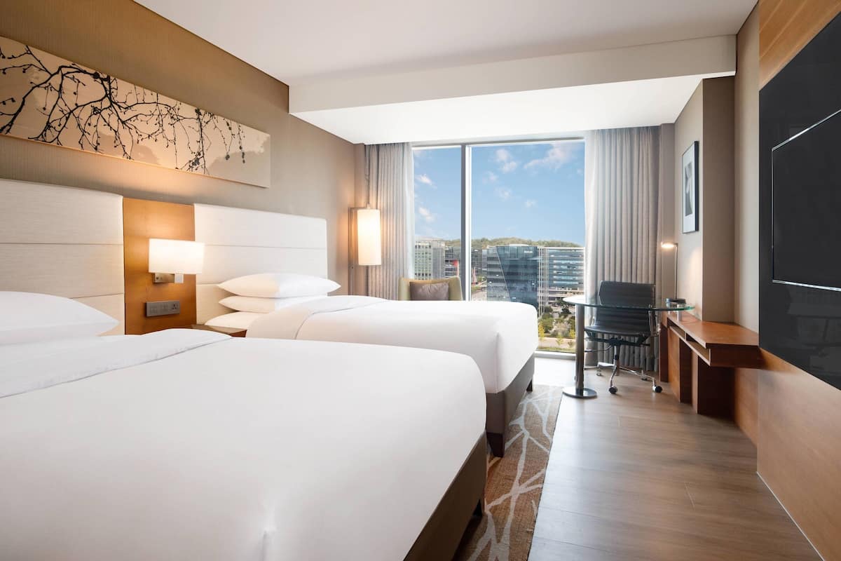 Quarto de hotel com duas camas, uma escrivaninha, uma cadeira, televisão e uma janela grande com vista para a cidade.