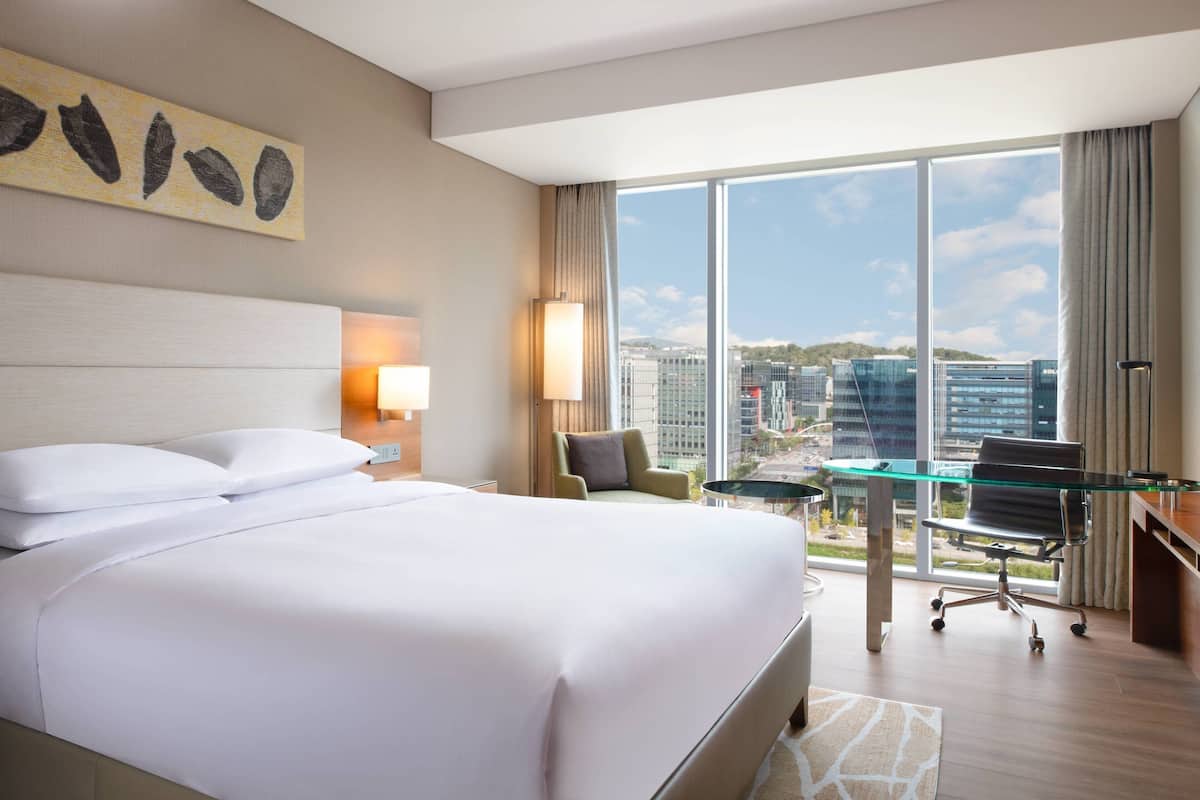 Quarto de hotel com uma cama grande, uma escrivaninha com tampo de vidro, uma cadeira e vista para a cidade através de grandes janelas.