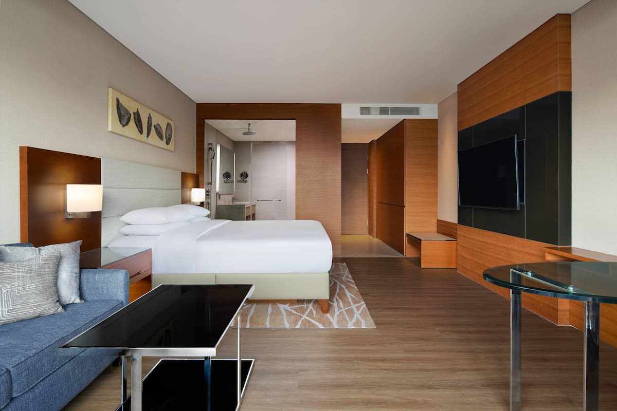 Quarto de hotel moderno com cama, sofá, mesa de vidro, televisão e banheiro visível através de uma porta aberta.