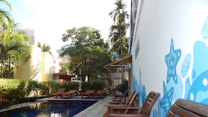Uma área na piscina com espreguiçadeiras de madeira, uma piscina pequena e um mural na parede.