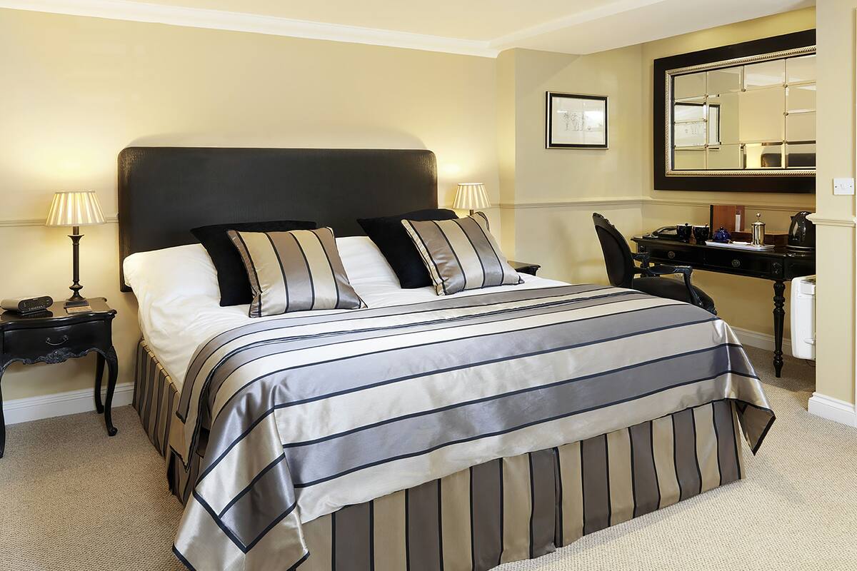 Uma cama bem arrumada com roupa de cama listrada, uma mesa de cabeceira com abajur, uma escrivaninha com espelho e uma cadeira.