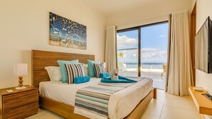 Um quarto espaçoso com uma cama grande, cabeceira de madeira, mesa de cabeceira com abajur e vista para a praia através de uma porta de vidro deslizante.