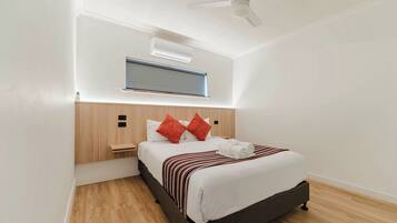 Quarto de hotel moderno com cabeceira de madeira, cama branca com almofadas vermelhas, manta listrada e ventoinha no teto.