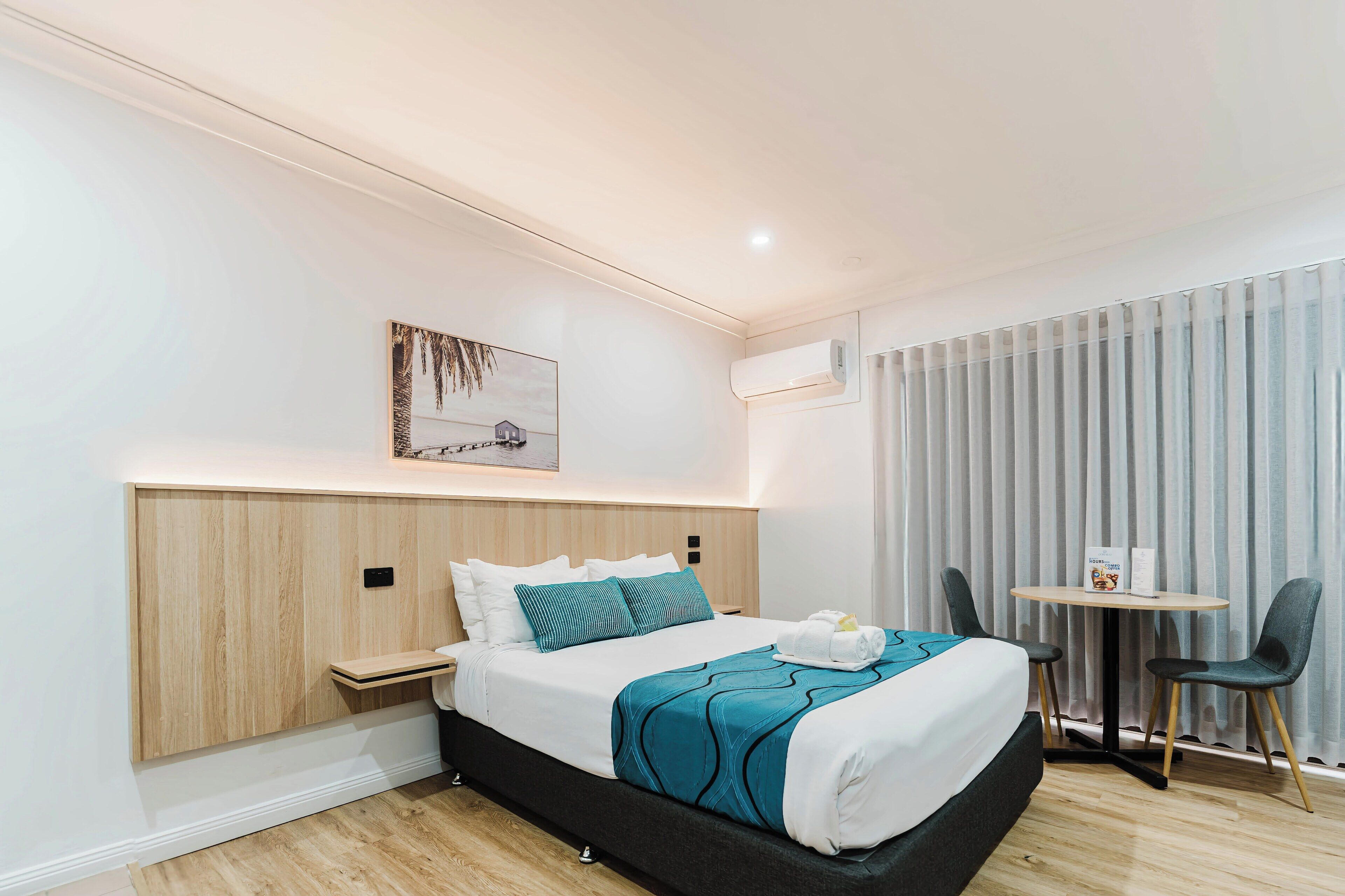 Quarto de hotel moderno com cama, mesa de cabeceira e uma pequena mesa redonda com cadeiras.