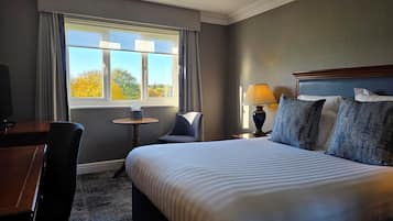 Quarto de hotel com uma cama grande, uma escrivaninha, uma cadeira, uma mesinha e um abajur.