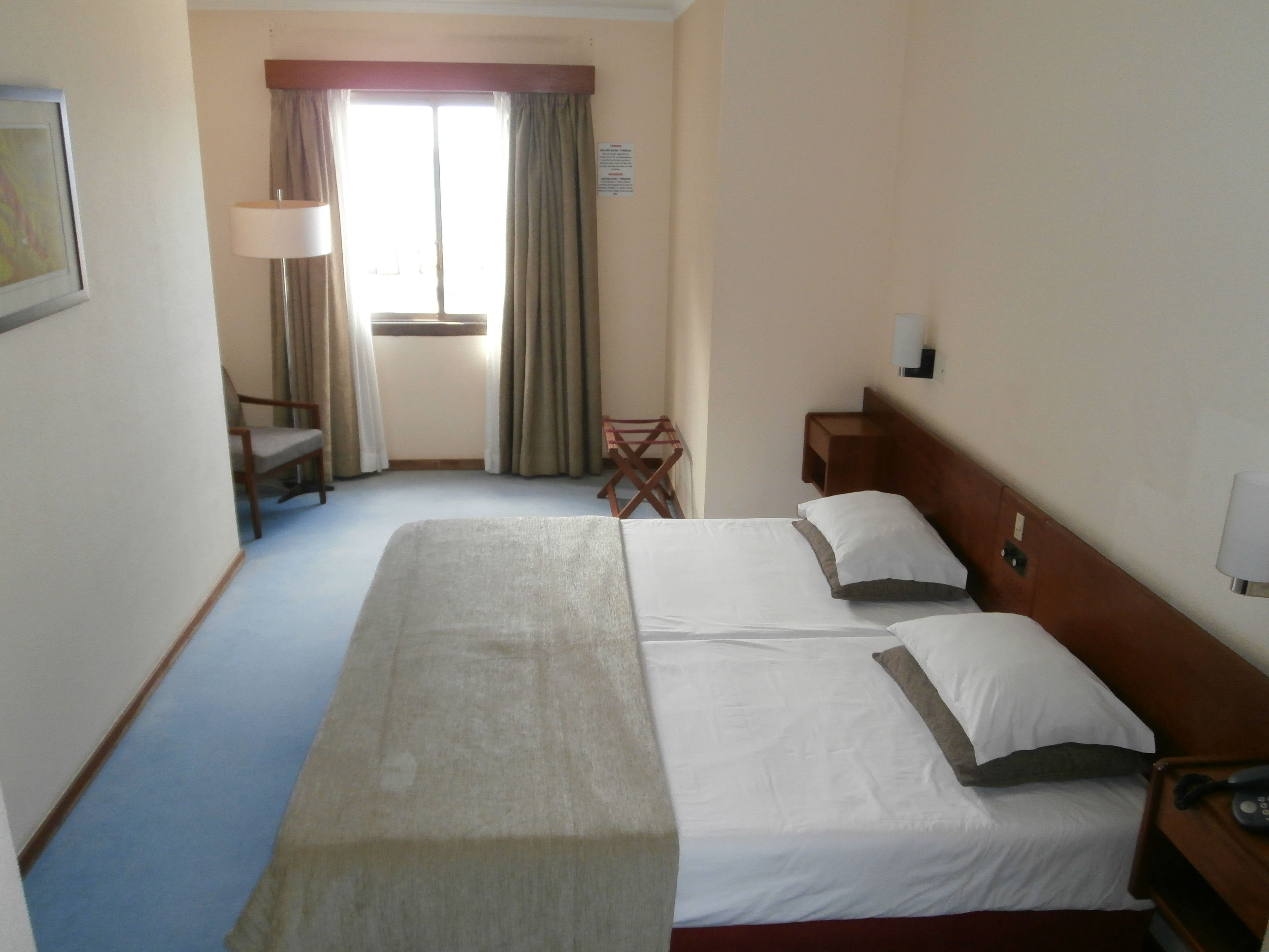 Quarto de hotel com uma cama grande, escrivaninha, cadeira e janela com cortinas.