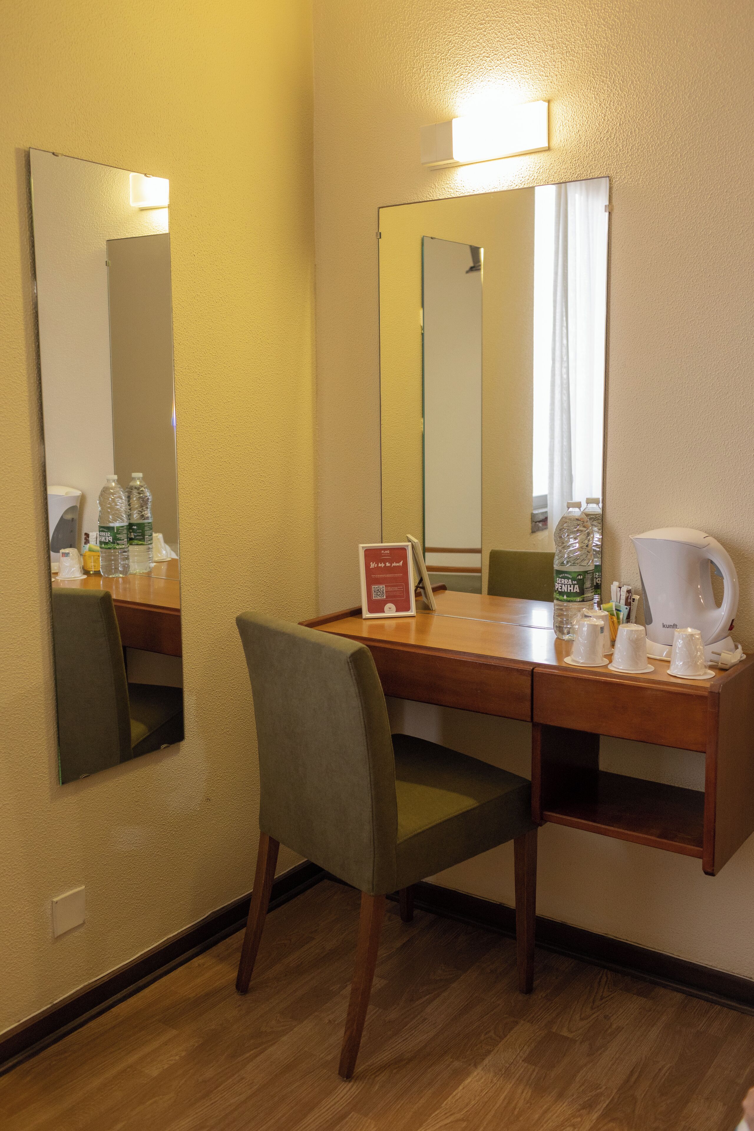 Quarto de hotel com escrivaninha de madeira, uma cadeira, um espelho, uma cafeteira e uma placa na parede.