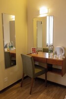 Quarto de hotel com escrivaninha de madeira, uma cadeira, um espelho, uma cafeteira e uma placa na parede.