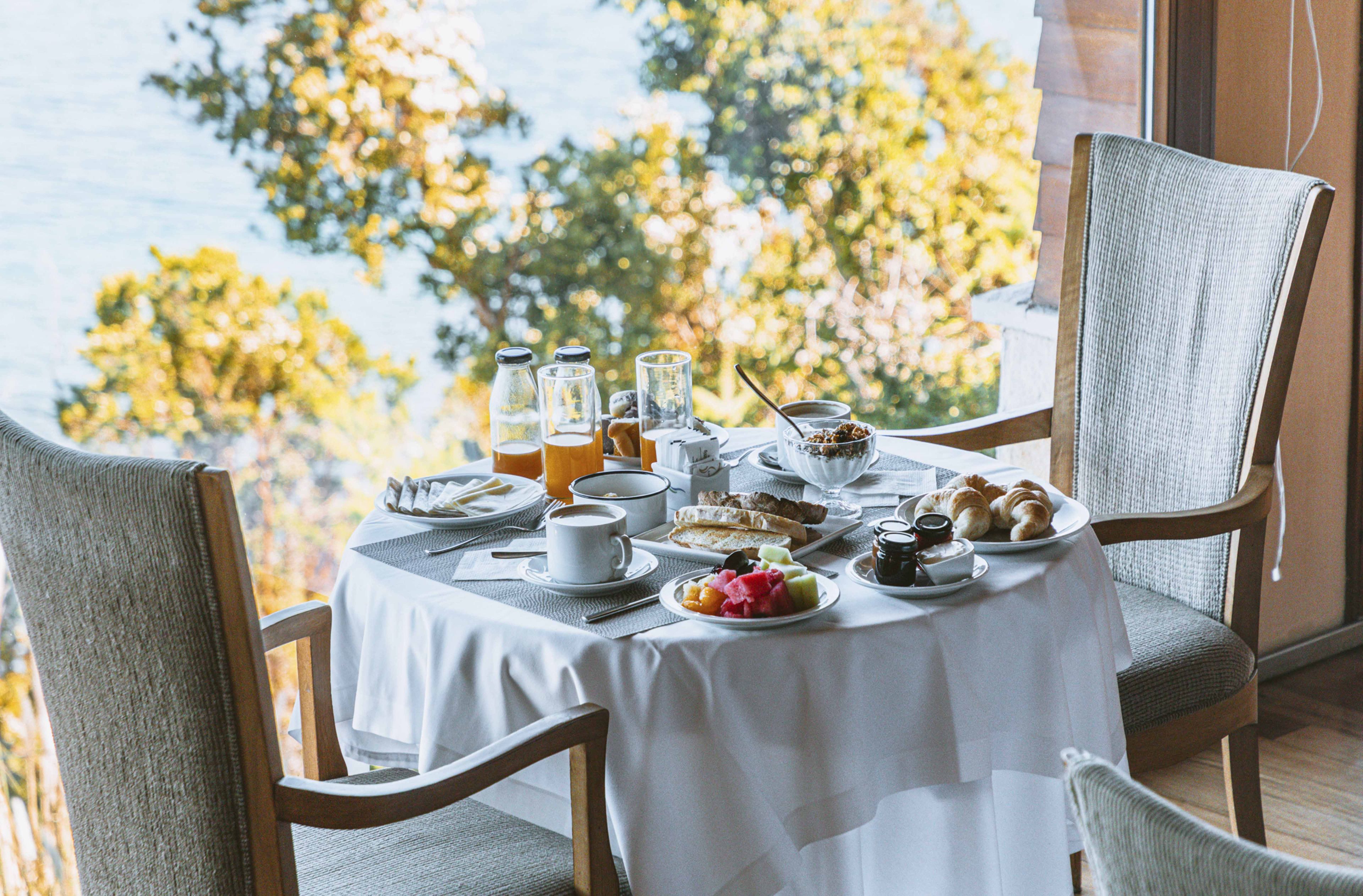 Mesa de café da manhã posta com diversos pratos, incluindo croissants, frutas e bebidas, com vista para uma paisagem cênica de árvores e um corpo d'água.