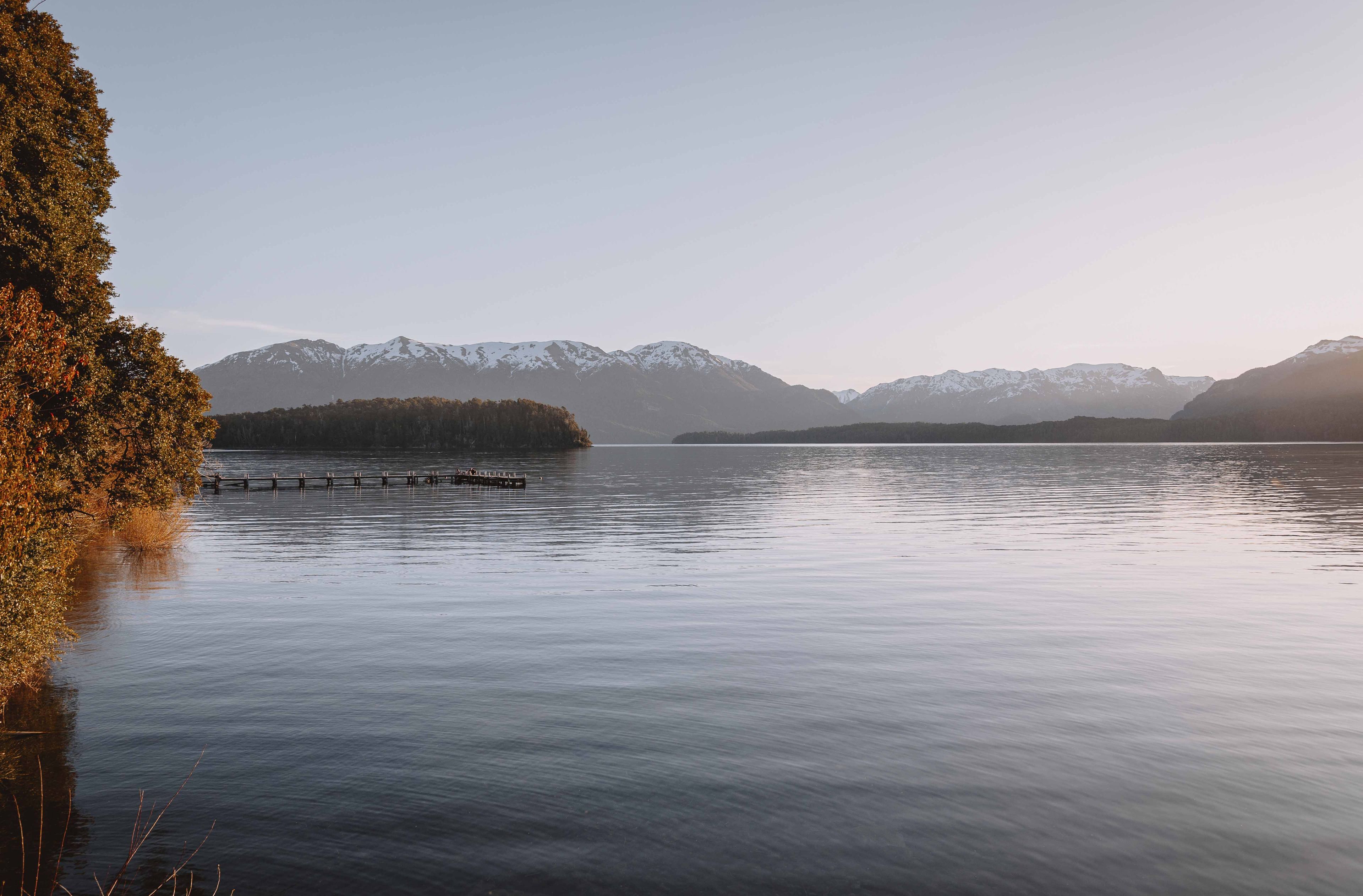 Um lago tranquilo com um cais que se estende sobre a água, rodeado por montanhas e árvores.