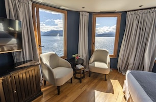 Quarto com cama, duas poltronas, uma mesa pequena com um vaso e uma TV em um armário. Janelas amplas oferecem vista para um lago e montanhas.