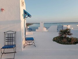 Um terraço à beira-mar com um edifício branco, uma pequena palmeira e duas cadeiras azuis.
