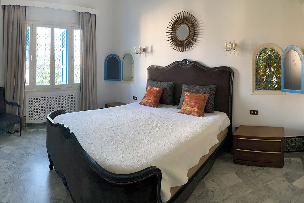 Quarto com cama grande, uma cadeira, mesa de cabeceira e janela com cortinas.