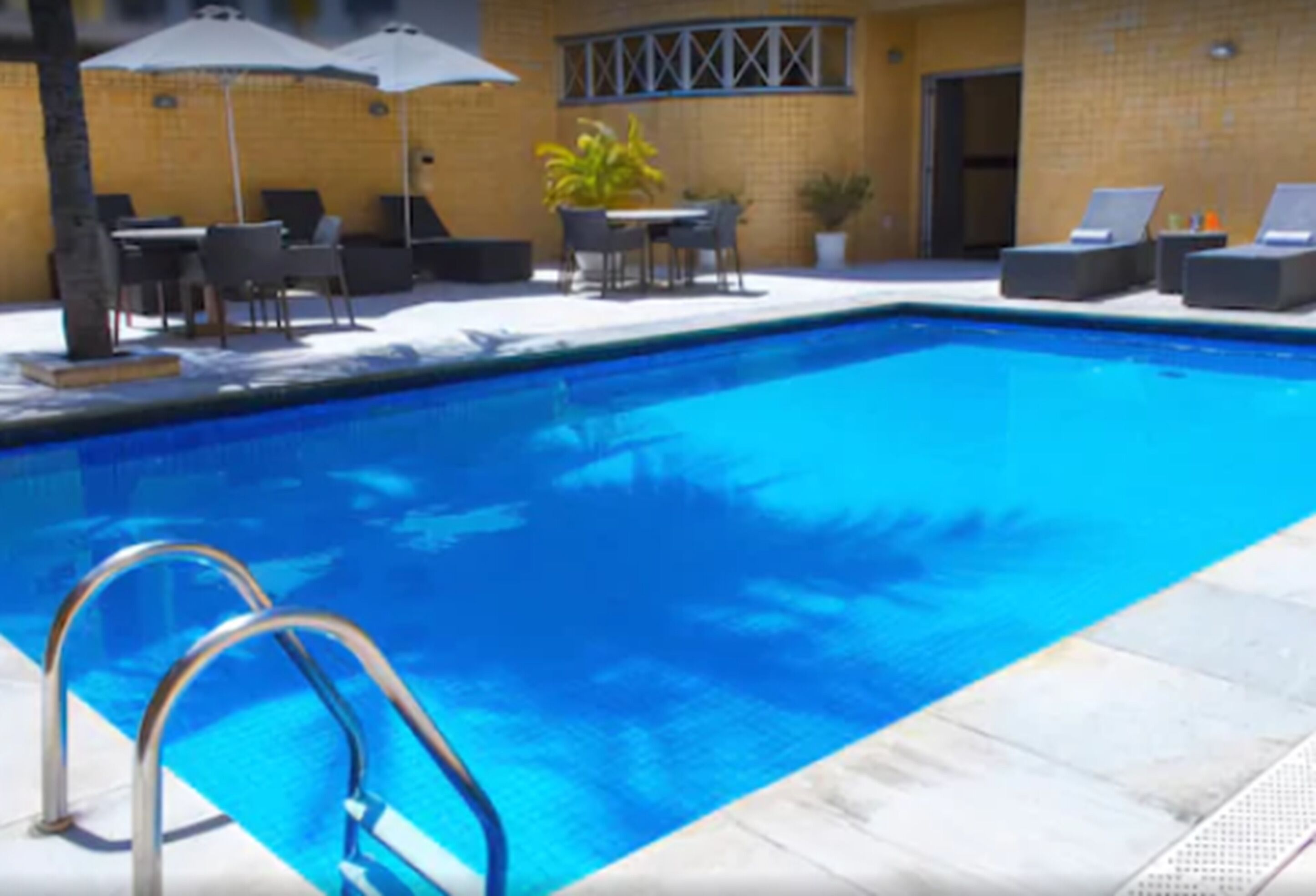 Piscina com escadas de metal, rodeada por espreguiçadeiras e guarda-sóis, com um muro de tijolos e uma porta ao fundo.