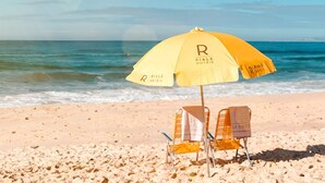 Uma praia com um guarda-sol amarelo e duas cadeiras com o logotipo "Rialle Hotels".