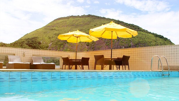 Uma piscina com espreguiçadeiras e guarda-sóis amarelos, rodeada por paredes revestidas e vista para uma colina verde.