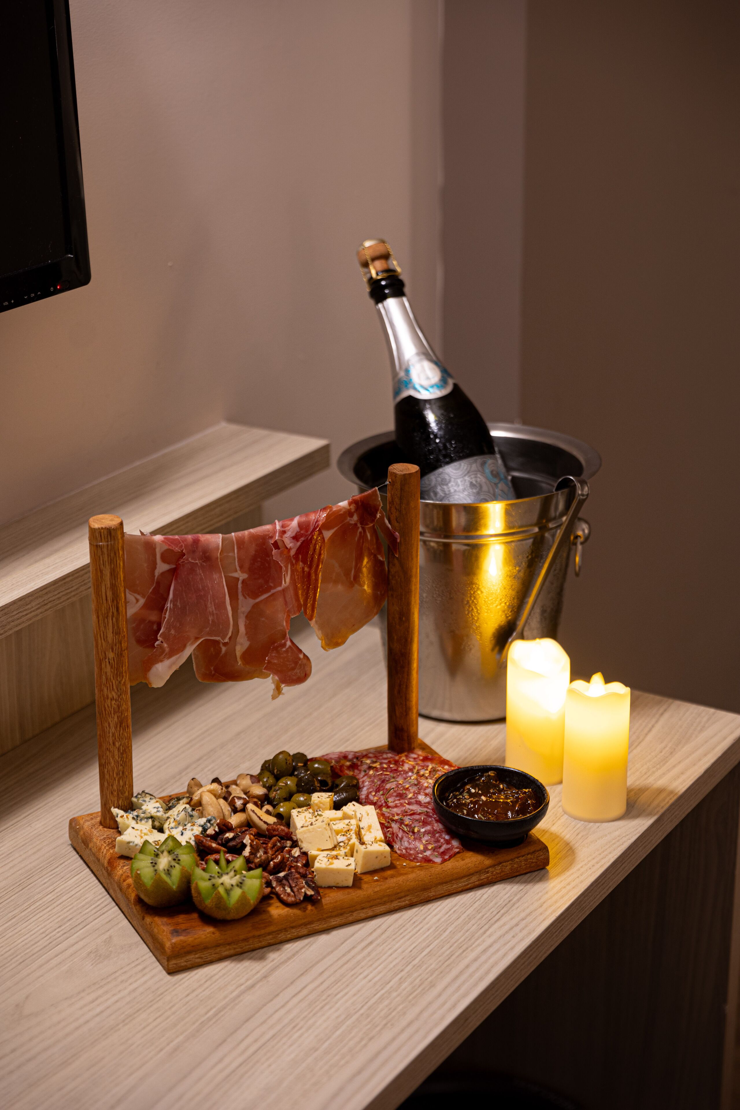 Uma tábua de madeira com diversos queijos, carnes e frutas, uma garrafa de champanhe em balde de metal e duas velas sobre uma superfície de madeira.