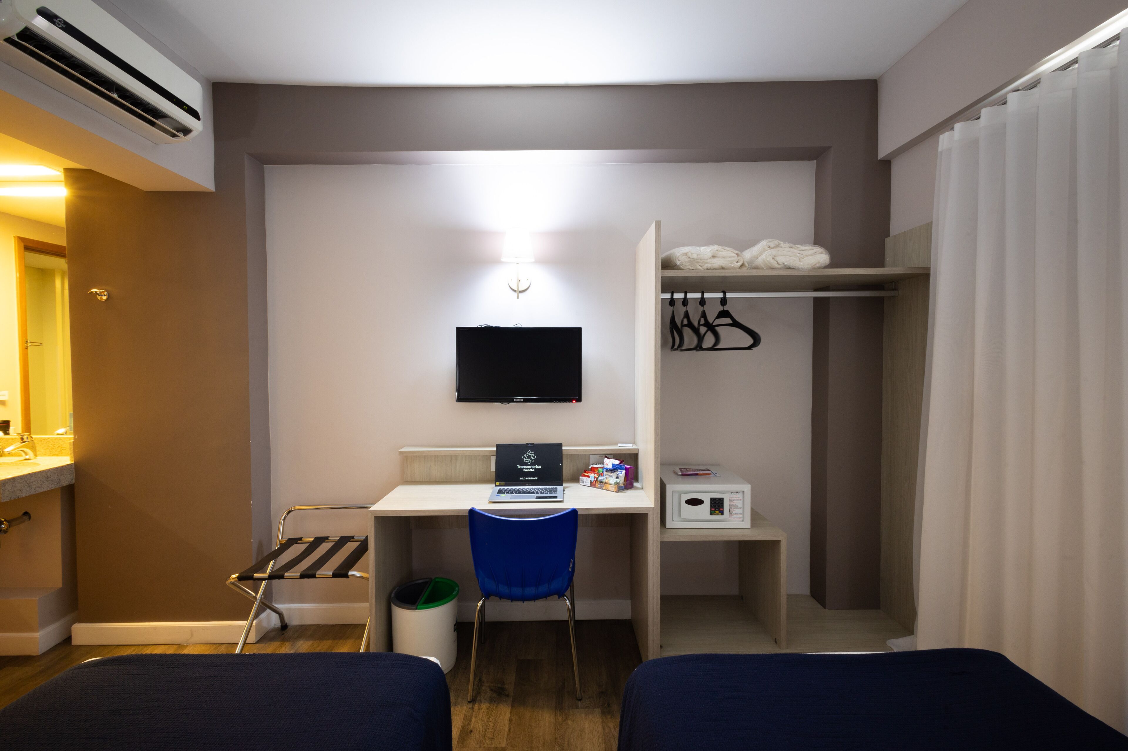 Quarto de hotel compacto com uma cama de solteiro, uma escrivaninha com cadeira, televisão, guarda-roupa e um pequeno cesto de lixo.