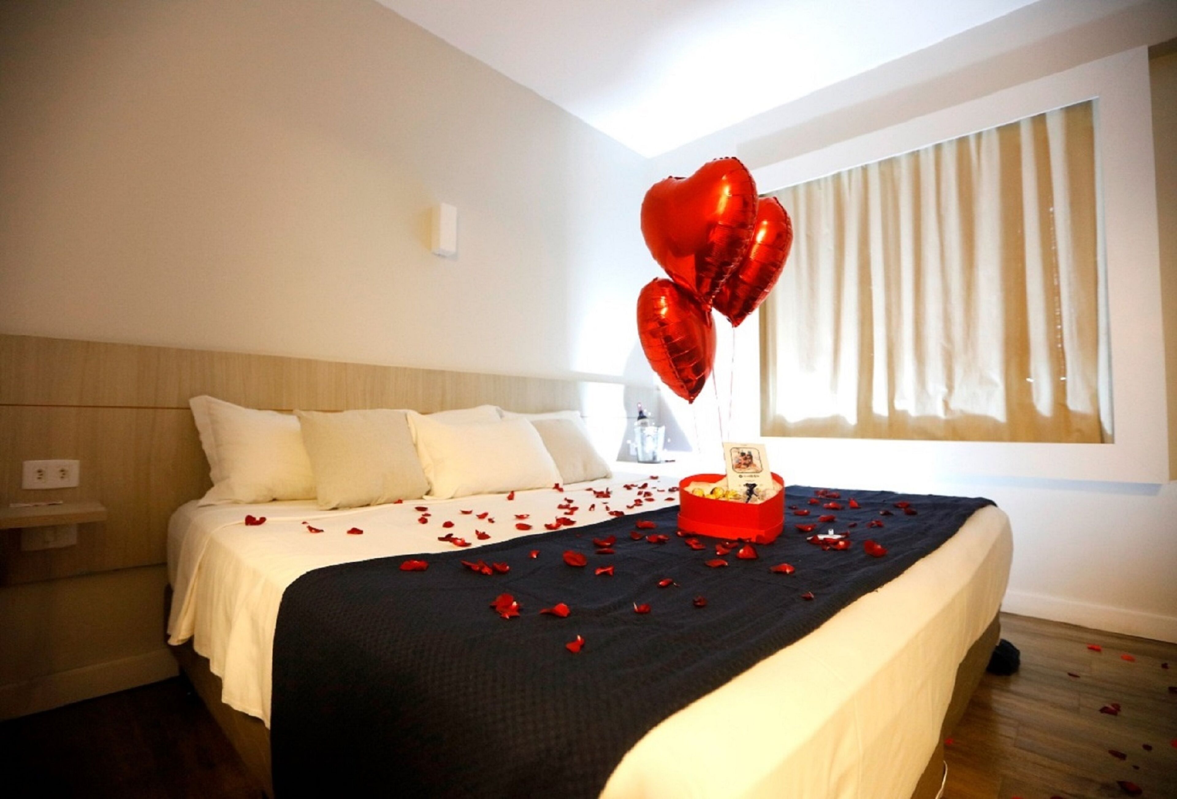 Quarto de hotel com cama bem arrumada, mesa de cabeceira e janela com cortinas.