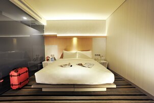 Quarto de hotel com cama, mala vermelha, mesa de cabeceira e abajur.